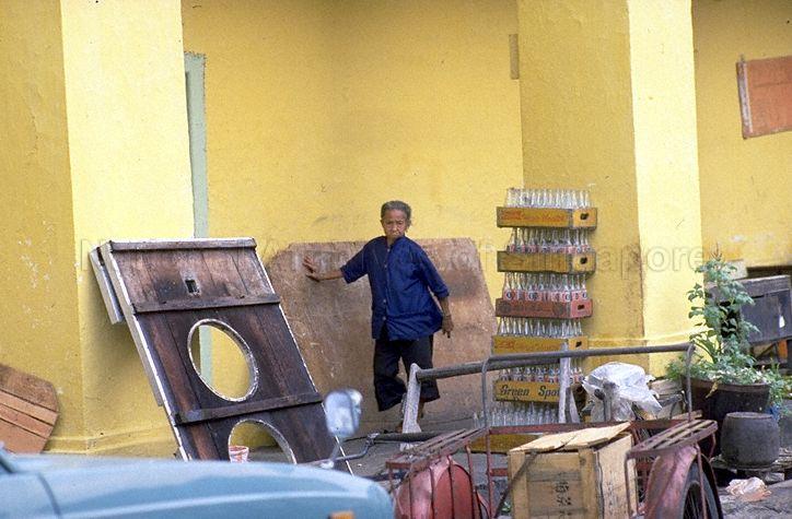 WOMAN IN TRADITIONAL SAM FOO WALKS PAST CRATES OF GREEN SPOT